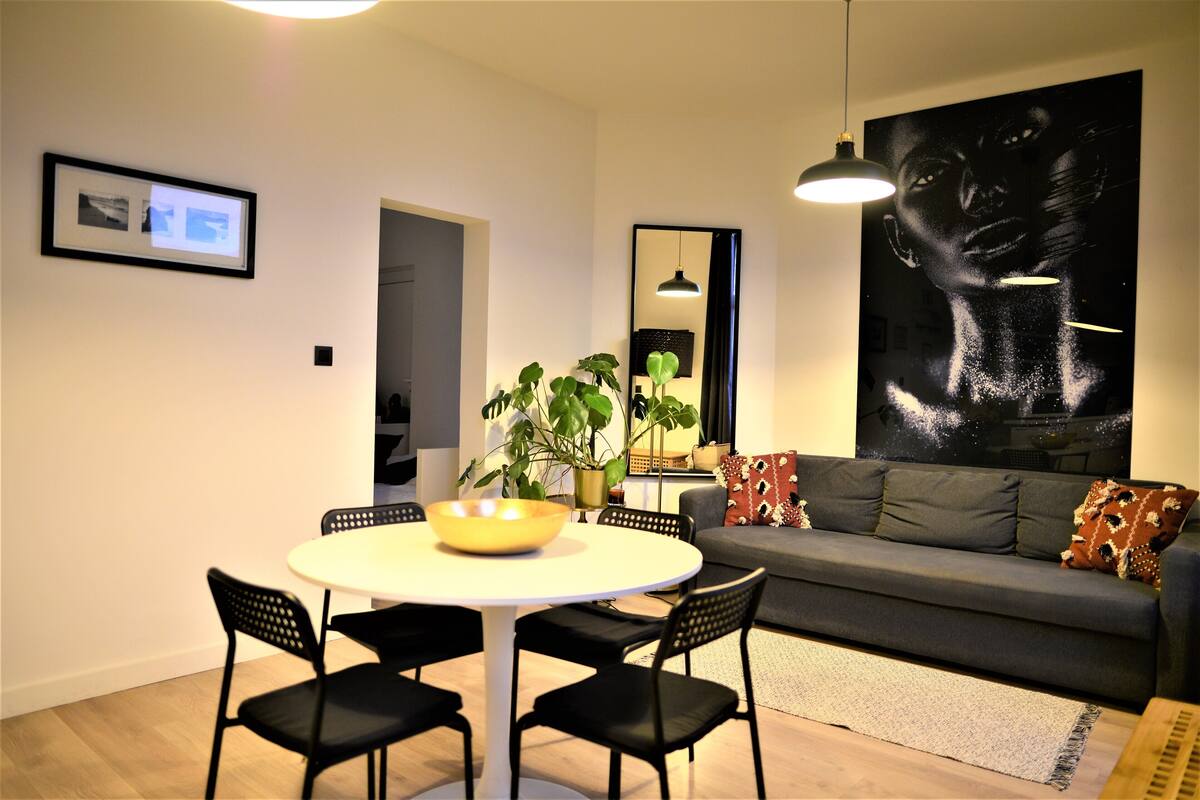 Uma sala de estar moderna com uma mesa de jantar redonda, cadeiras pretas, um sofá cinza com almofadas estampadas, uma grande pintura abstrata e um vaso com planta.