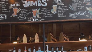 Uma área de bar com um cardápio em lousa exibindo diversos drinques, uma pessoa segurando uma cerveja, e prateleiras com garrafas e copos.