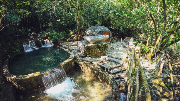 Um ambiente natural com uma fonte de pedra, um caminho de pedra e uma estrutura em forma de cúpula.