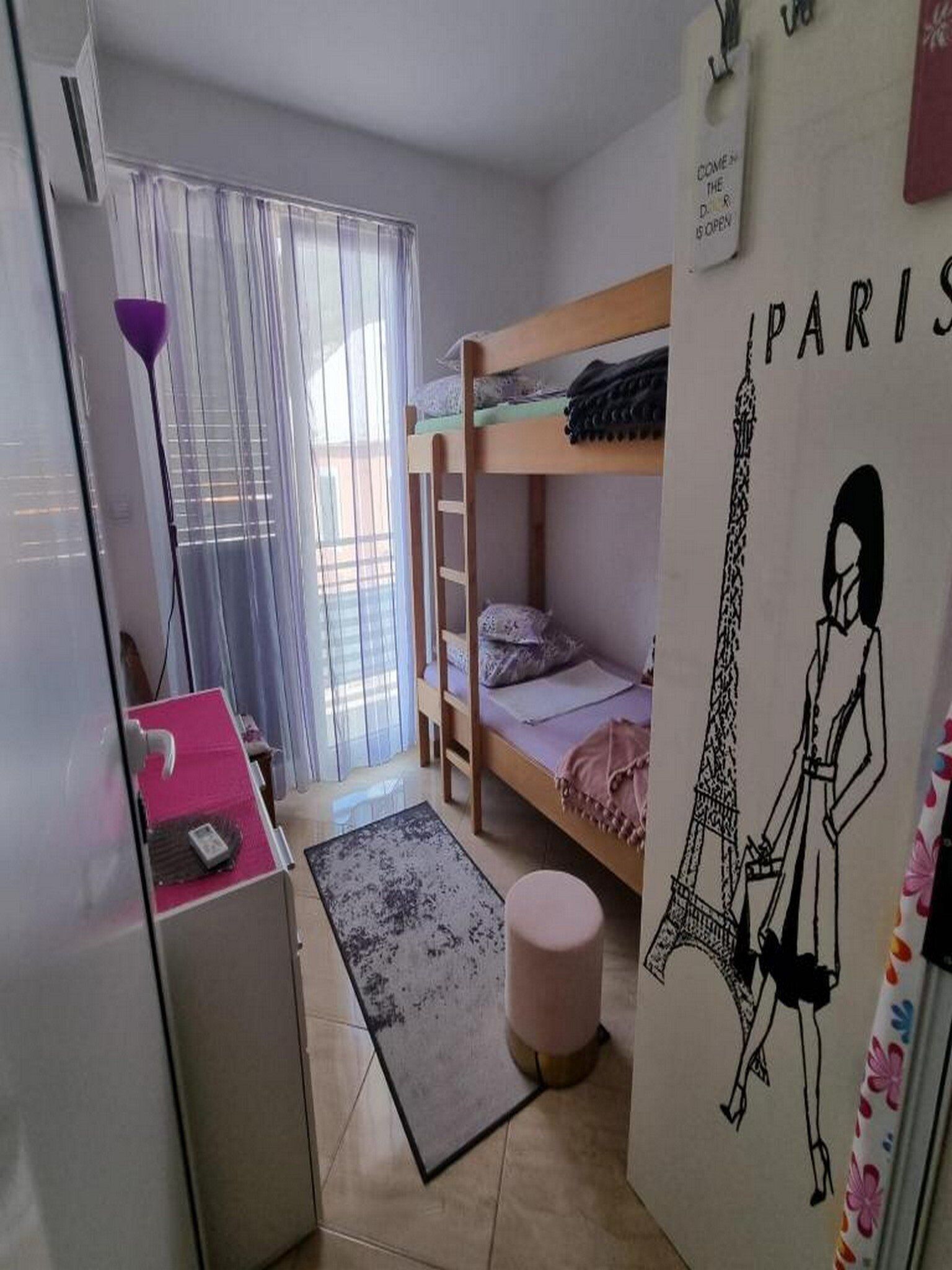 Quarto com beliches, uma escrivaninha, uma cadeira, um banquinho pequeno, um pôster da Torre Eiffel e uma janela com cortinas.