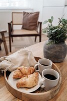 Uma bandeja de madeira com croissants e xícaras de café sobre uma mesa, uma cadeira e um vaso com planta.