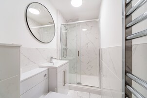Uma casa de banho moderna com chuveiro em cabine de vidro, espelho redondo e parede revestida a mármore branco.