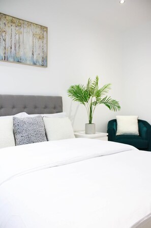 Quarto com cama, cabeceira cinza, poltrona verde, mesa de cabeceira branca, planta em vaso e um quadro na parede.