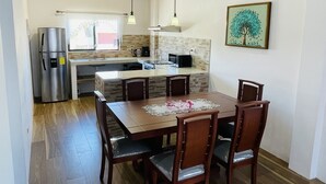 Cozinha compacta com geladeira, micro-ondas e fogão. Há uma mesa de jantar com cadeiras e uma janela com cortinas.