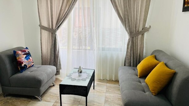 Uma sala de estar com dois sofás cinza, uma mesa de centro preta pequena e uma janela com cortinas leves.
