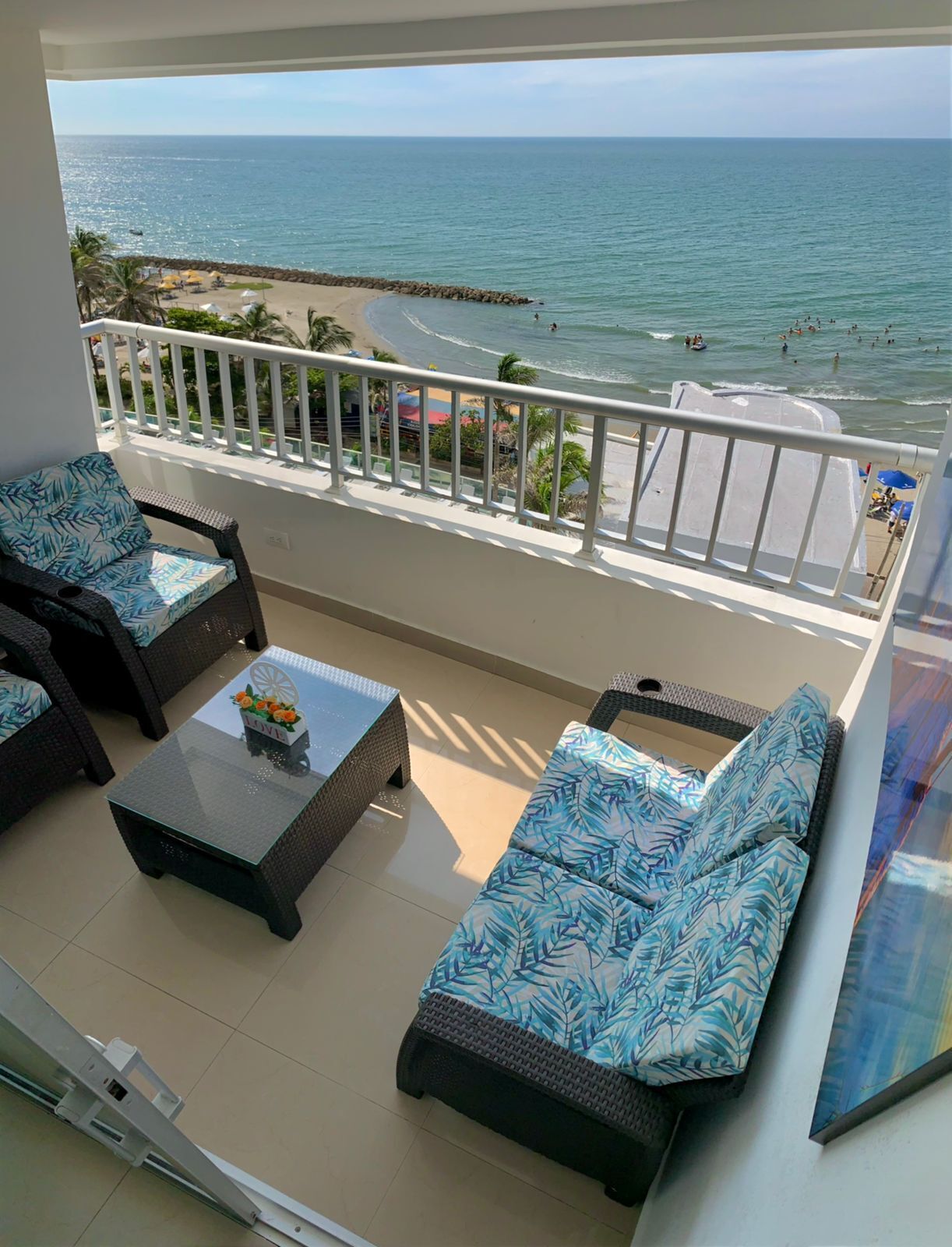 Uma varanda com mobiliário de vime, uma mesa de centro de vidro e vista para a praia e o oceano.