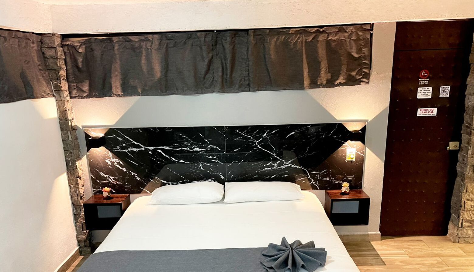 Quarto de hotel com uma cama grande, cabeceira de mármore, duas mesas de cabeceira e porta com fechadura de segurança.