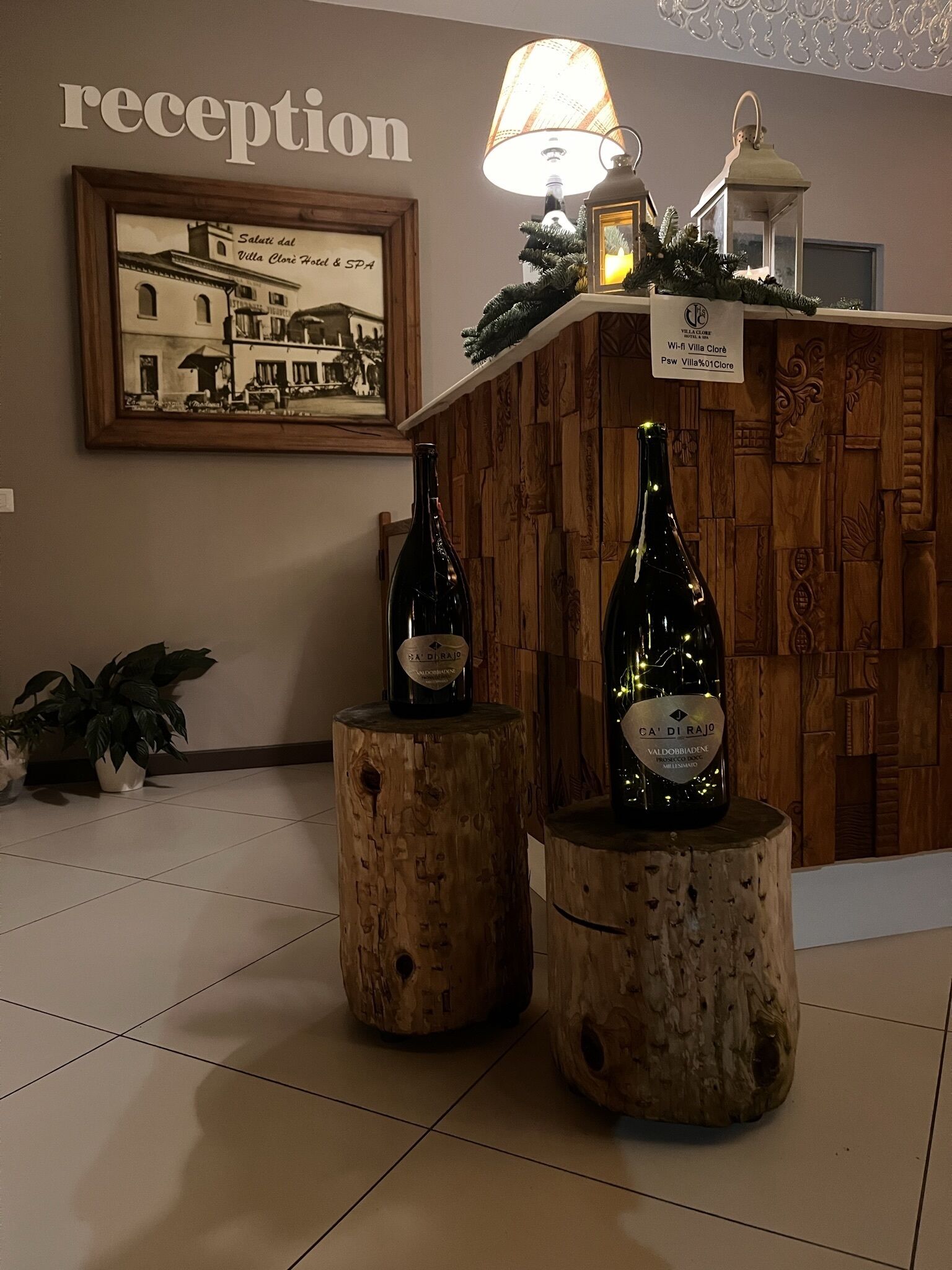 Uma área de recepção com um quadro de boas-vindas, dois banquinhos de madeira com garrafas de vinho, um abajur e um sinal indicando disponibilidade de Wi-Fi.