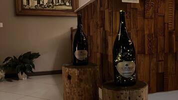 Uma área de recepção com um quadro de boas-vindas, dois banquinhos de madeira com garrafas de vinho, um abajur e um sinal indicando disponibilidade de Wi-Fi.