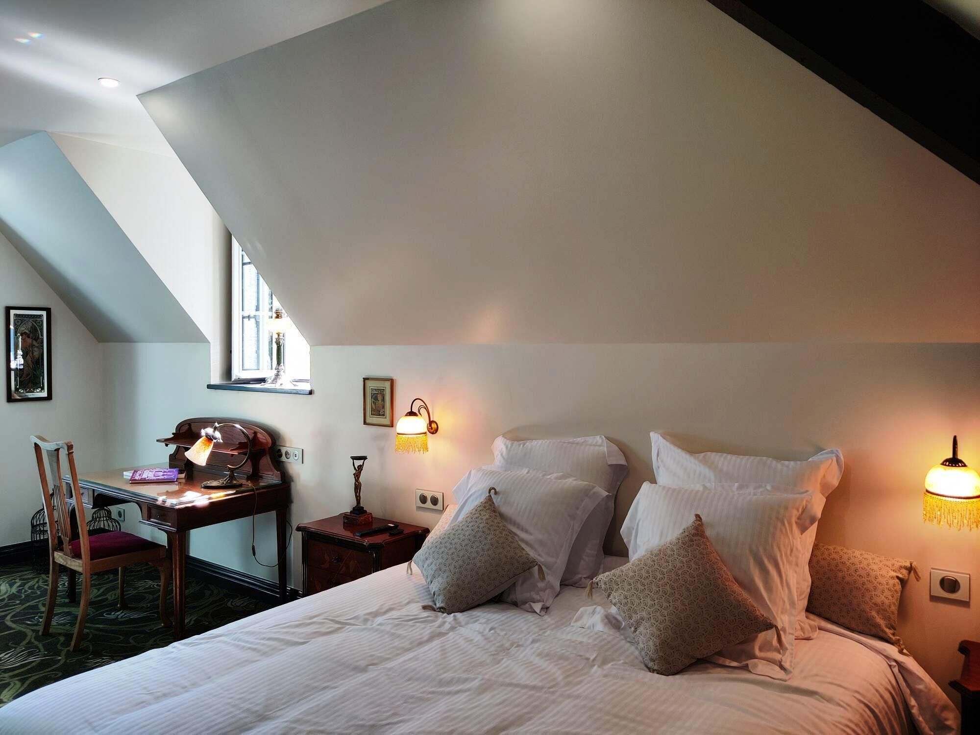 Um quarto com teto inclinado, uma escrivaninha com abajur, uma cadeira e uma cama com vários travesseiros.