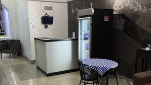 Cozinha pequena e moderna com frigorífico, micro-ondas e pia. Há uma mesa com toalha xadrez e duas cadeiras. O chão é em mosaico e as paredes são brancas.