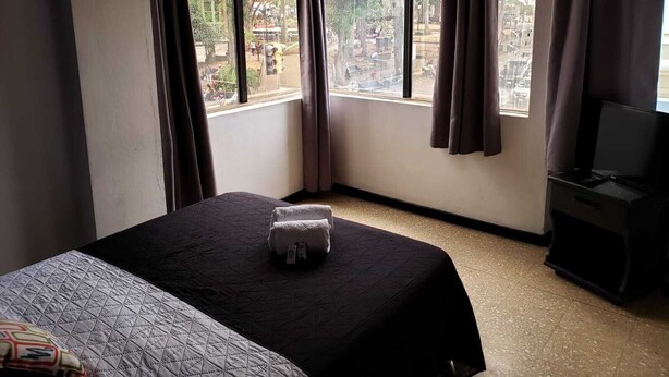 Quarto de hotel com cama, televisão e janelas amplas com cortinas.