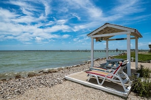 Um gazebo à beira-mar com mesa e cadeiras, com vista para um mar calmo e um píer ao longe.
