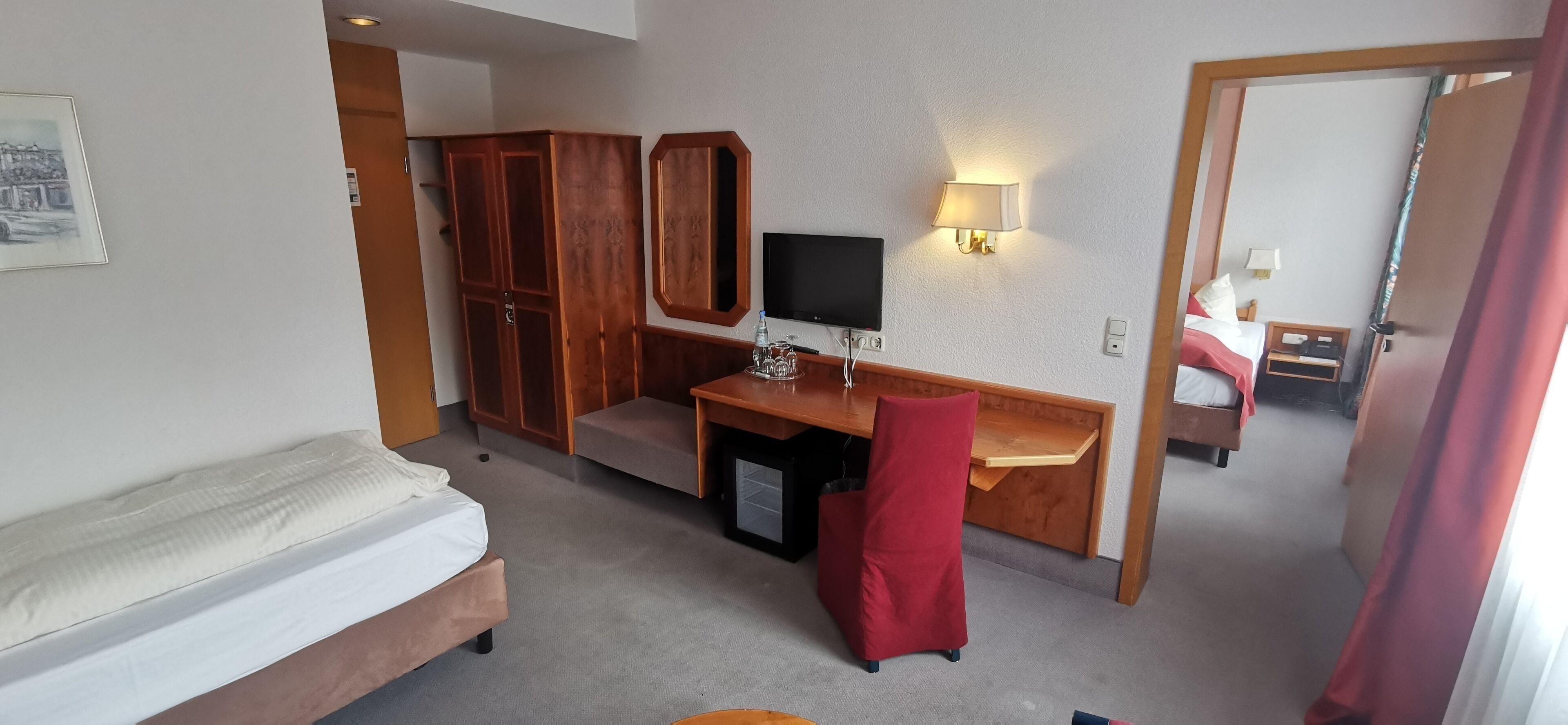 Quarto de hotel com cama, escrivaninha, cadeira, televisão e guarda-roupa.