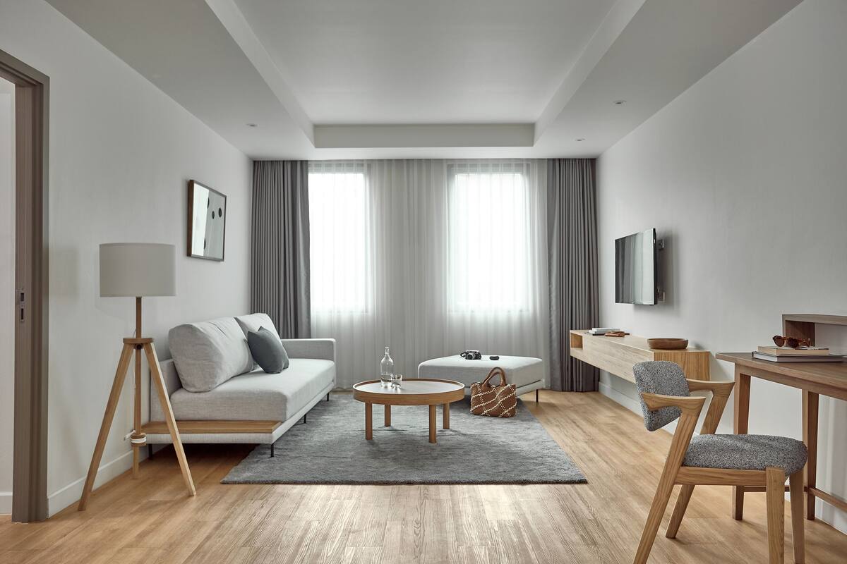 Uma sala de estar moderna com sofá, mesa de centro e poltronas, uma luminária, televisão e uma grande janela com cortinas.