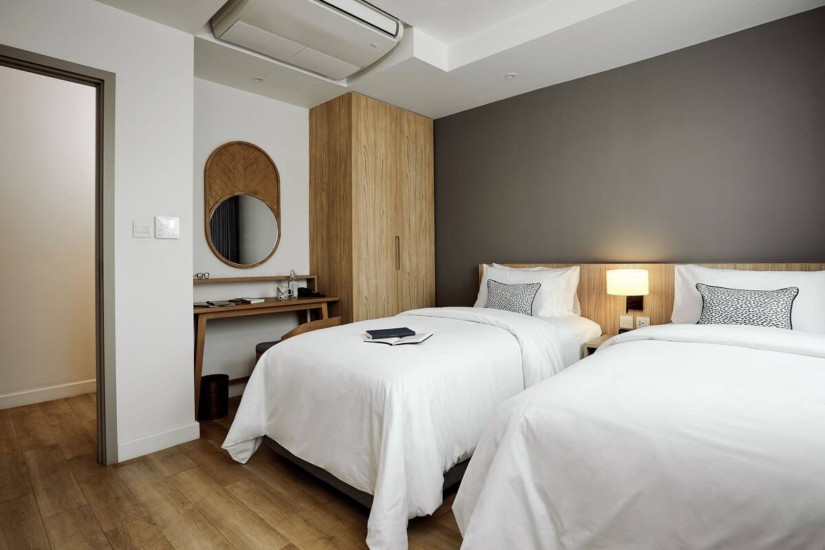 Quarto de hotel com duas camas, um guarda-roupa de madeira, um espelho redondo e uma mesa de cabeceira com abajur.