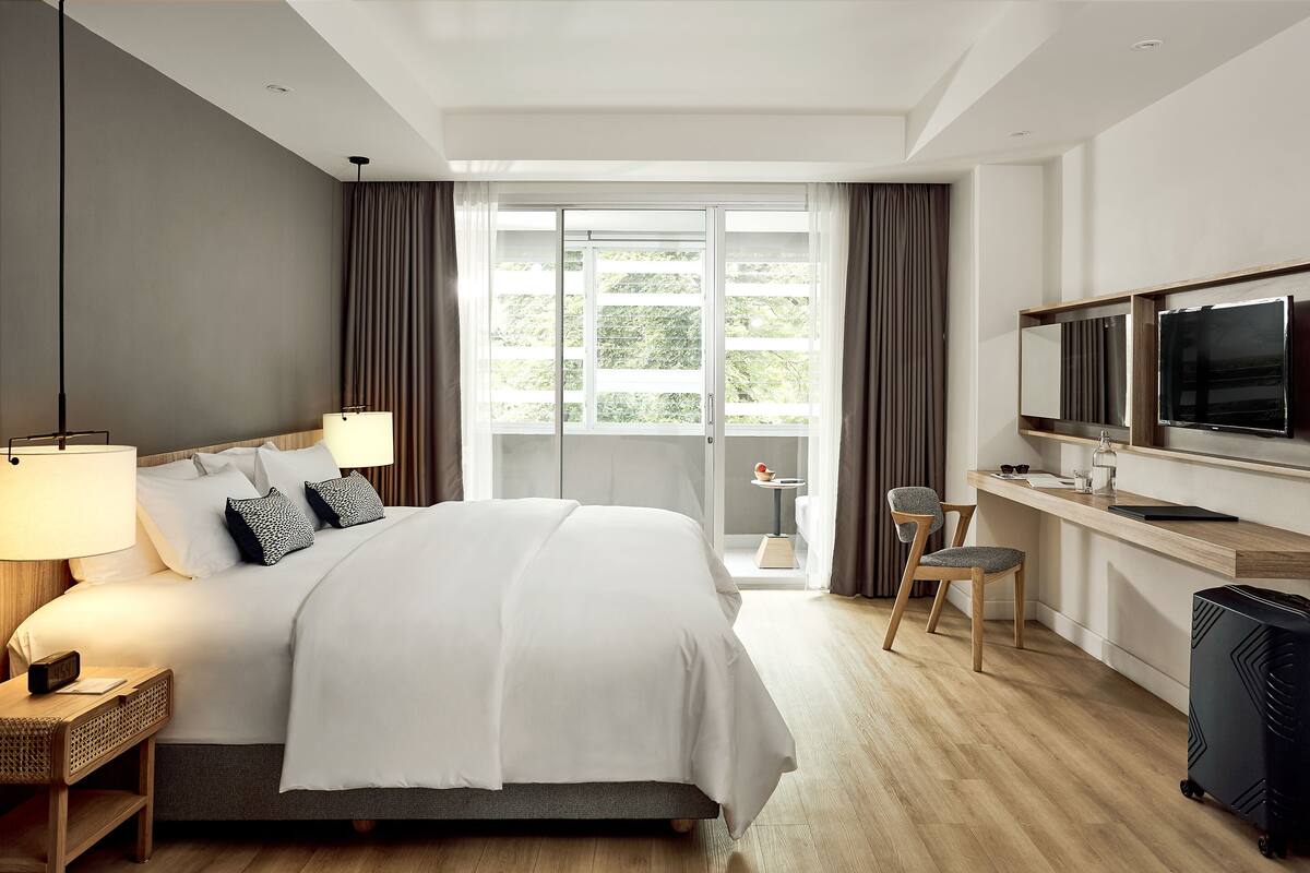 Quarto de hotel moderno com uma cama grande, abajures, uma escrivaninha com cadeira, televisão e vista para o verde lá fora.