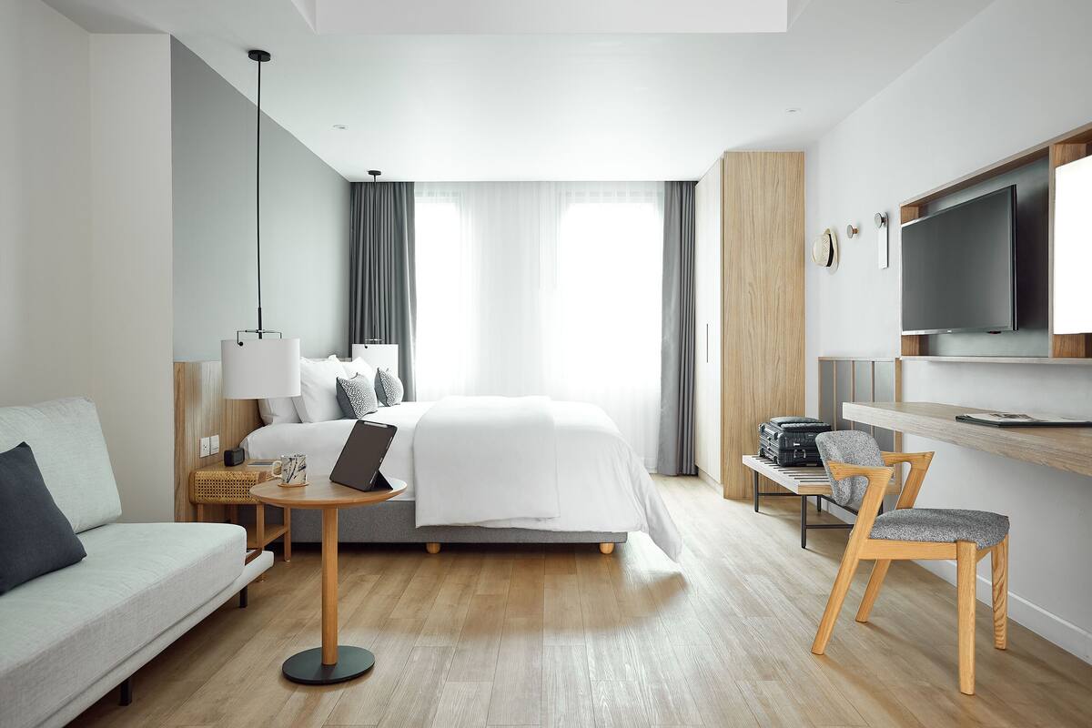 Quarto de hotel moderno com cama, sofá, escrivaninha com laptop, cadeira, televisão e janela com cortinas.