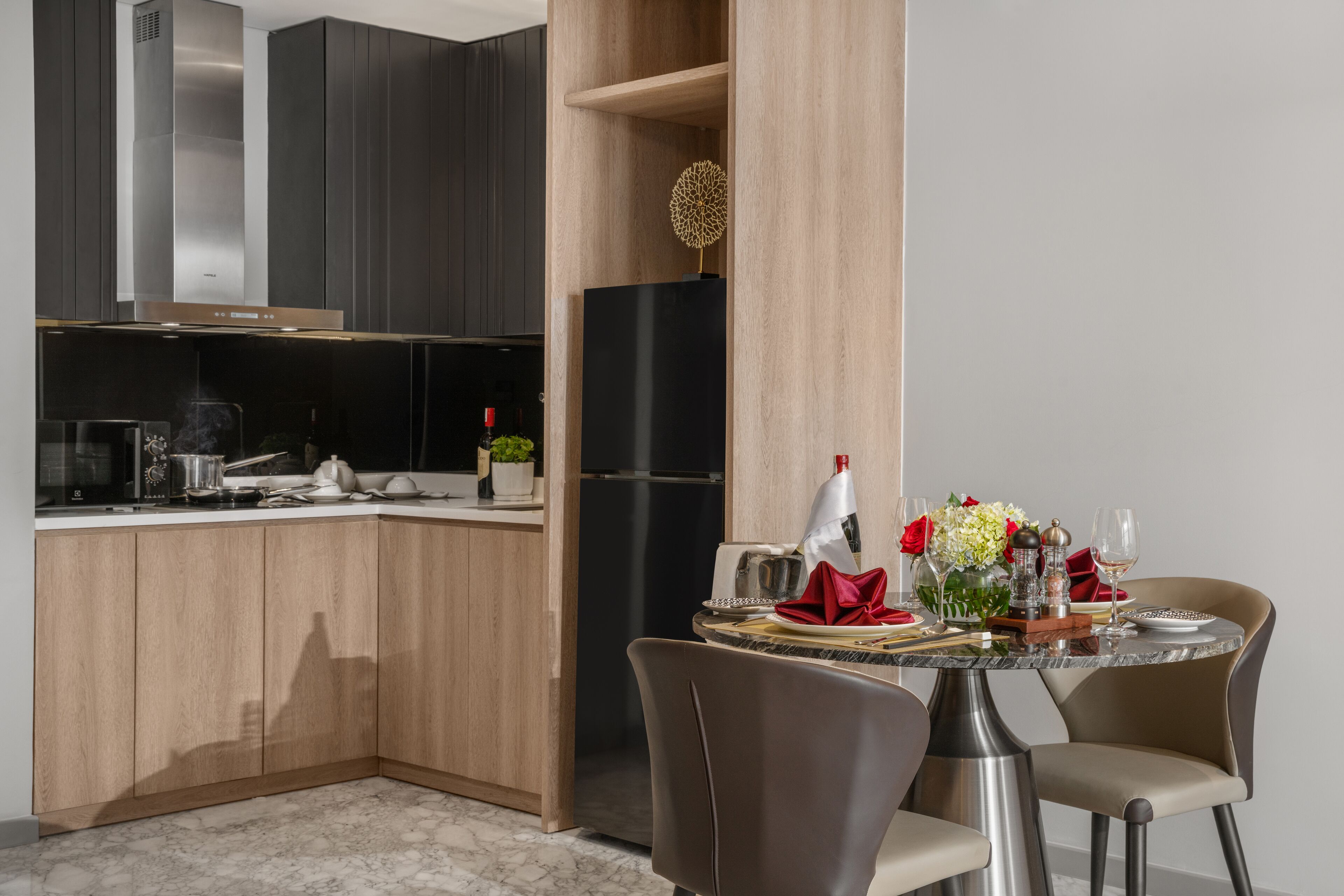 Uma cozinha moderna com armários escuros, um exaustor de aço inoxidável e uma área de jantar com mesa de mármore e duas cadeiras.