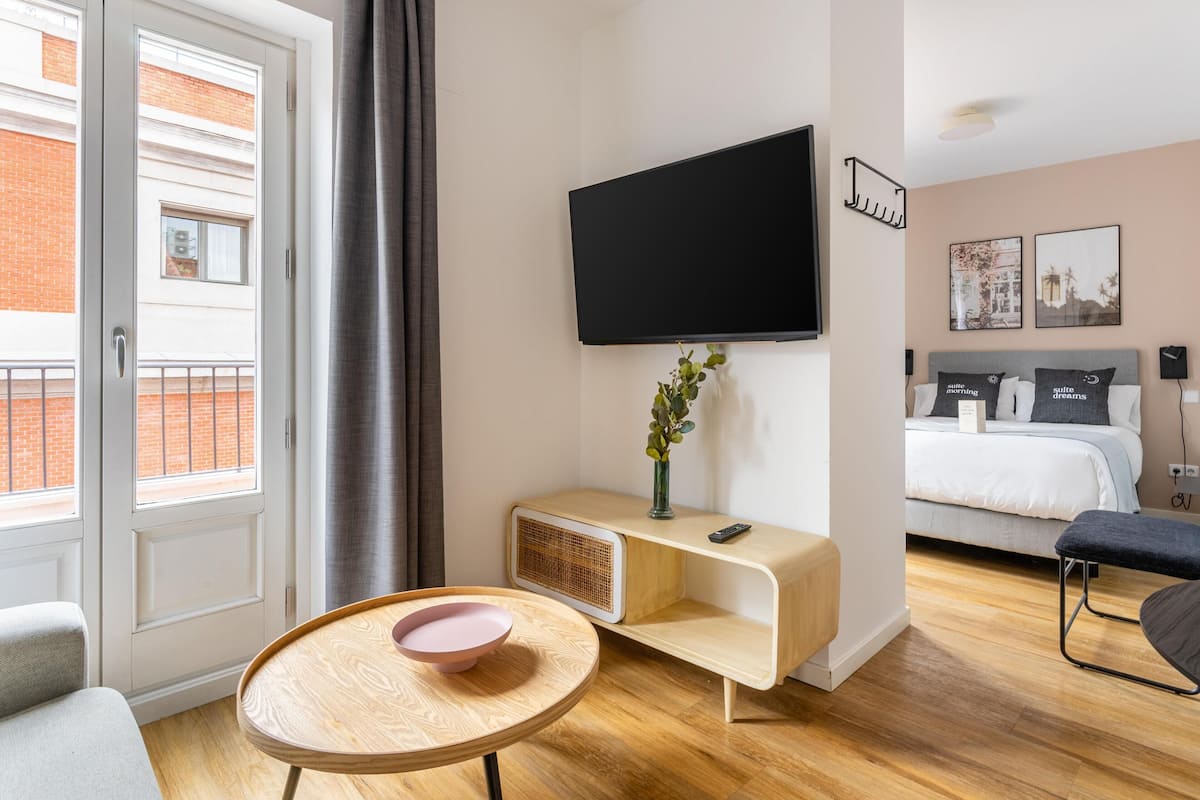Quarto de hotel moderno com uma televisão de tela plana grande, uma cama com colcha branca, uma pequena mesa redonda de madeira e uma tigela rosa, além de uma janela com vista para um prédio de tijolos.