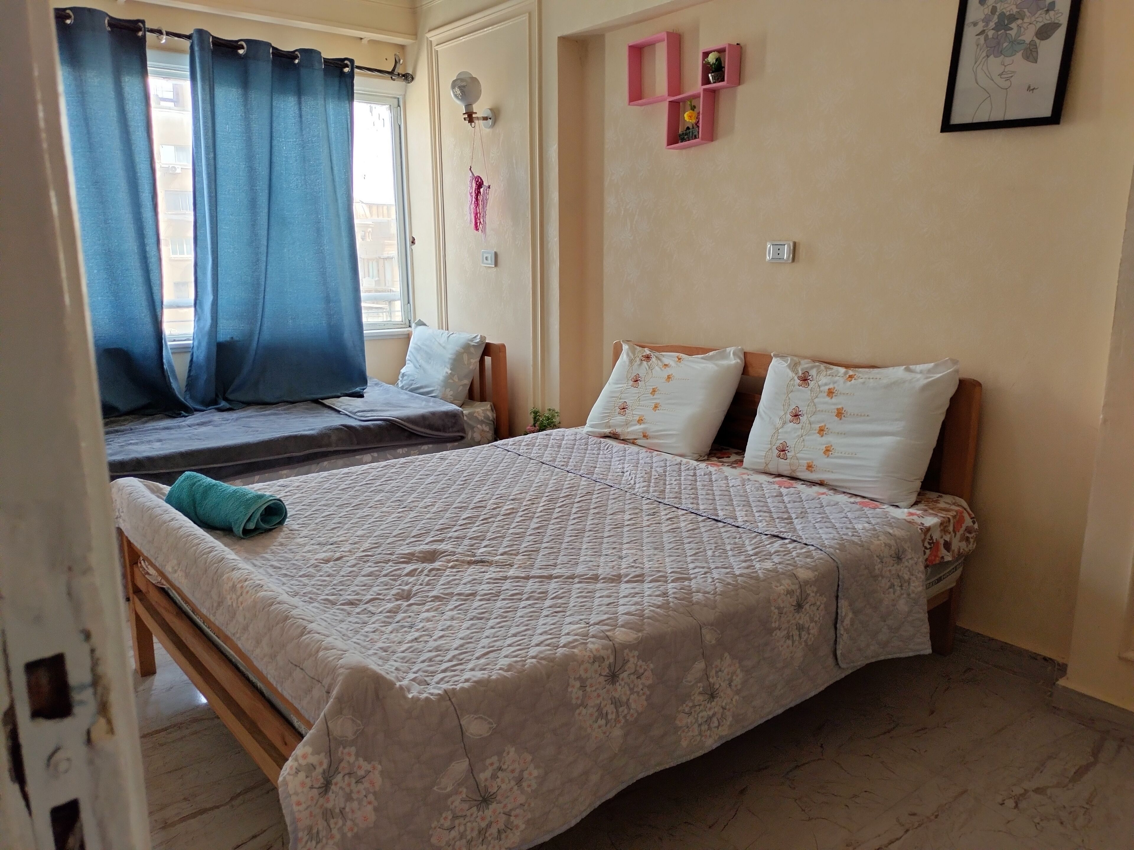 Quarto com duas camas, janela com cortinas azuis, luminária fixada na parede e quadro emoldurado.