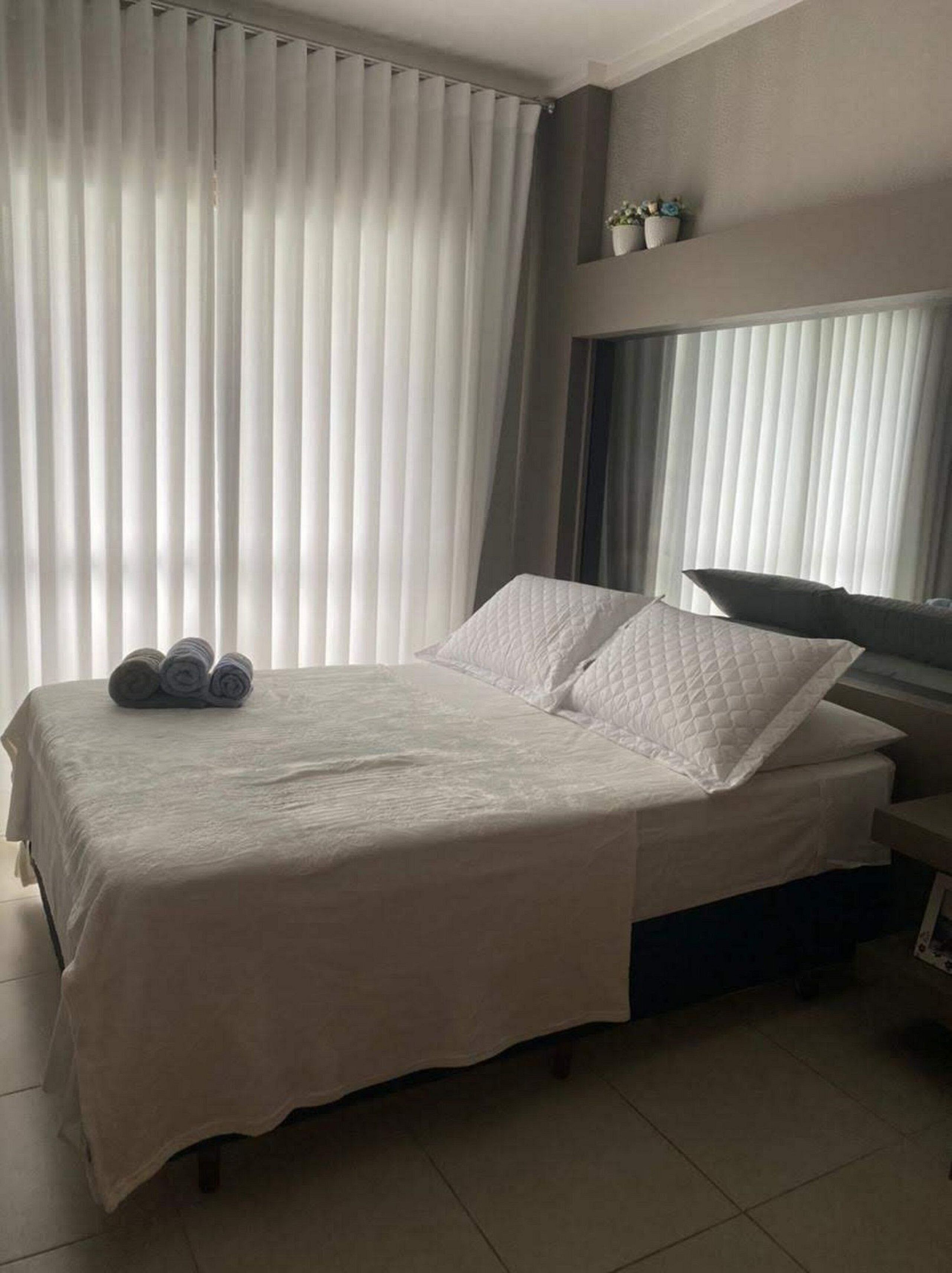 Cama bem arrumada com travesseiros e toalhas enroladas, mesa de cabeceira com abajur e janela com persianas verticais.