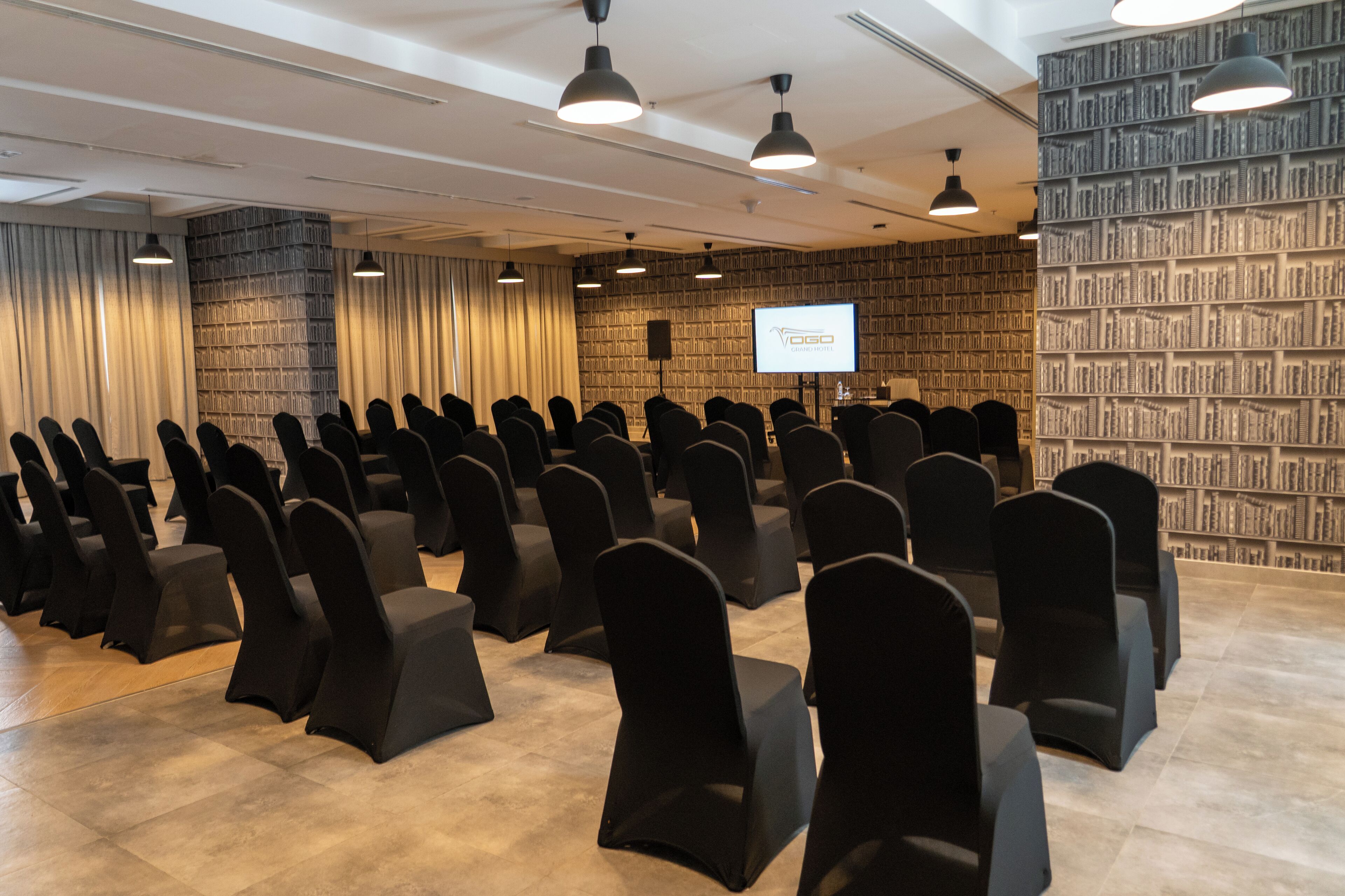 Uma sala de conferência com fileiras de cadeiras, uma tela de projeção e uma parede de estantes.