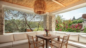 Uma área de refeições com mesa e cadeiras de madeira, teto de fibra trançada e vista para árvores e flores.