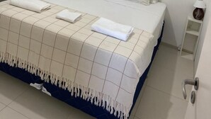 Uma cama bem arrumada com colcha xadrez e travesseiros brancos, um enfeite na parede e uma mesinha de cabeceira com uma luminária.
