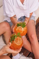 Duas pessoas segurando copos com bebidas e fatias de cítricos e folhas de hortelã.