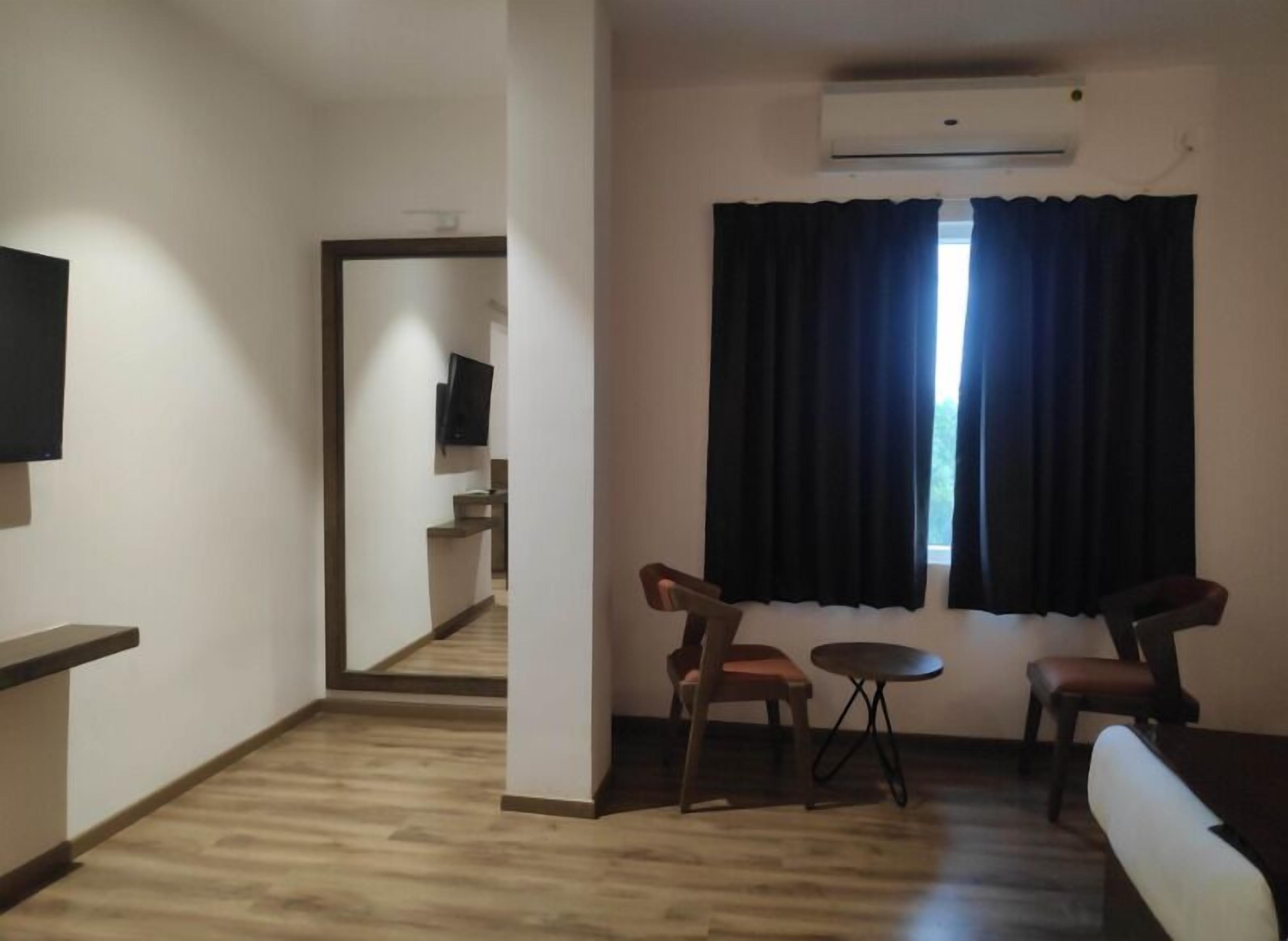 Quarto com piso de madeira, televisão de tela plana na parede, uma pequena mesa redonda, duas cadeiras e uma janela com cortinas escuras.