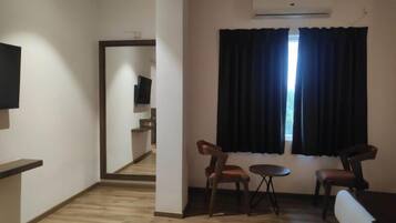 Quarto com piso de madeira, televisão de tela plana na parede, uma pequena mesa redonda, duas cadeiras e uma janela com cortinas escuras.