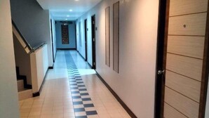Um corredor de hotel com chão xadrez, uma porta de madeira à direita e uma escadaria à esquerda.