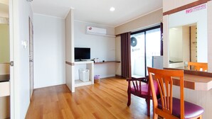 Quarto de hotel com piso de madeira, televisão em um suporte, uma cadeira, uma mesa pequena e uma janela com cortinas.