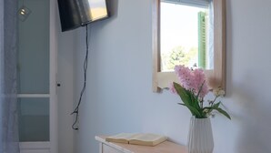Quarto com escrivaninha de madeira, vaso branco com flores, televisão na parede e janela com vista para o verde.
