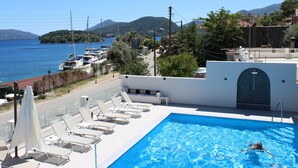 Uma piscina com espreguiçadeiras, vista para o mar e barcos ao fundo.