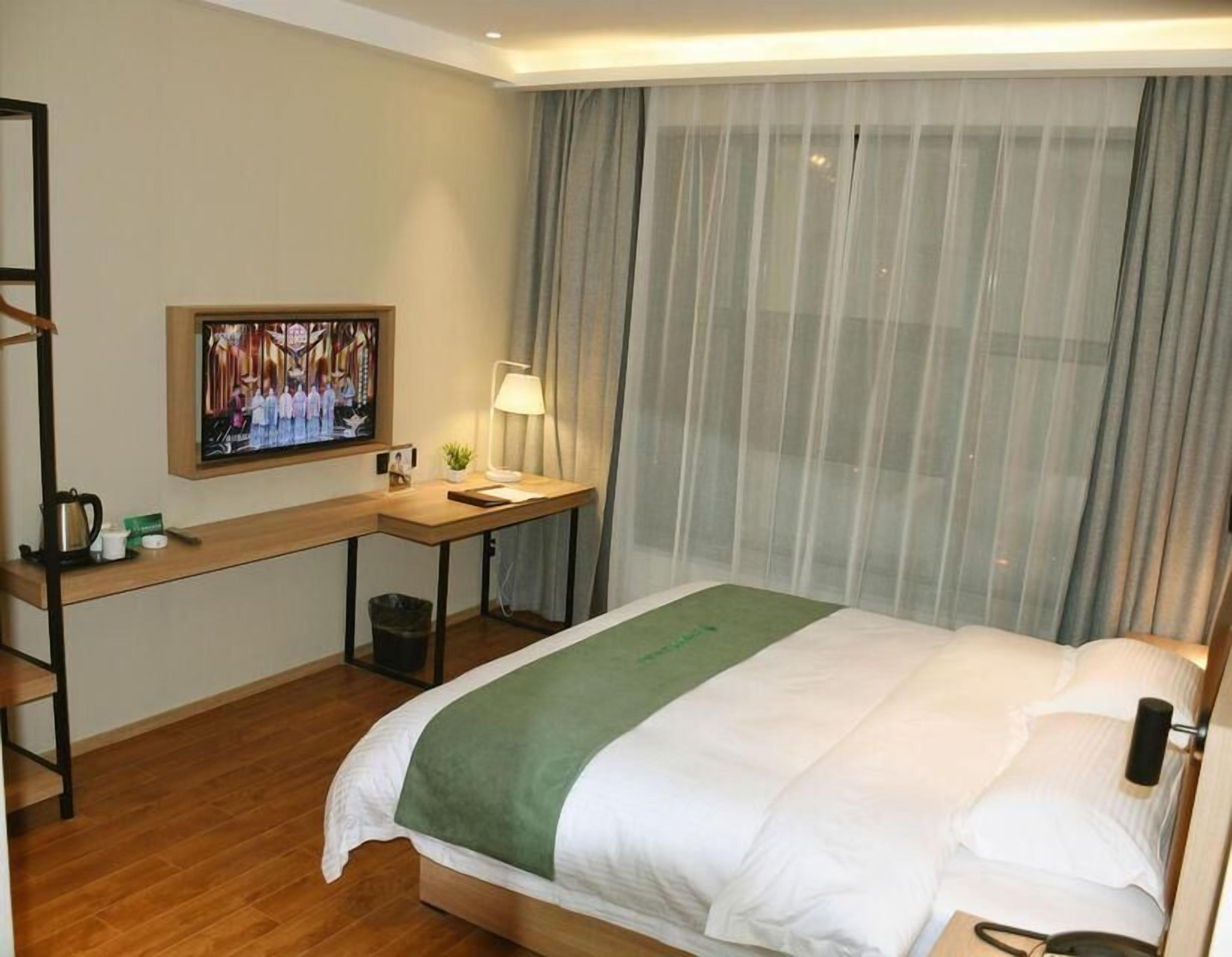 Quarto de hotel com cama, escrivaninha, televisão, luminária e janela com cortinas.