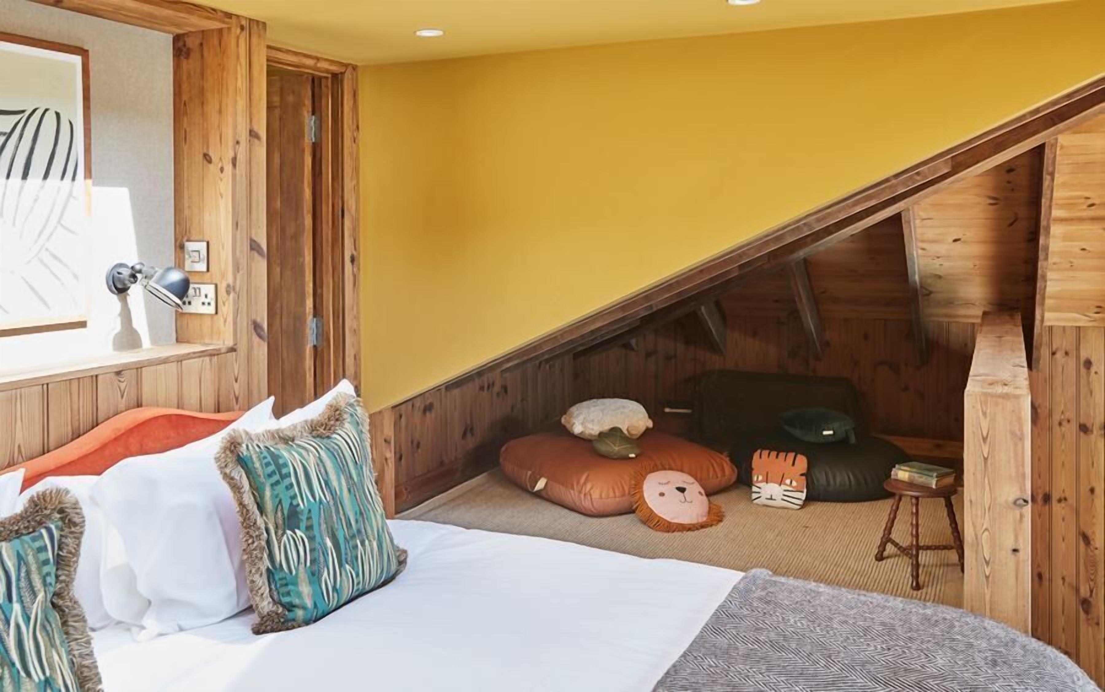 Um quarto de sótão aconchegante com teto inclinado, uma cama com vários travesseiros, um banquinho pequeno e uma luminária na parede.