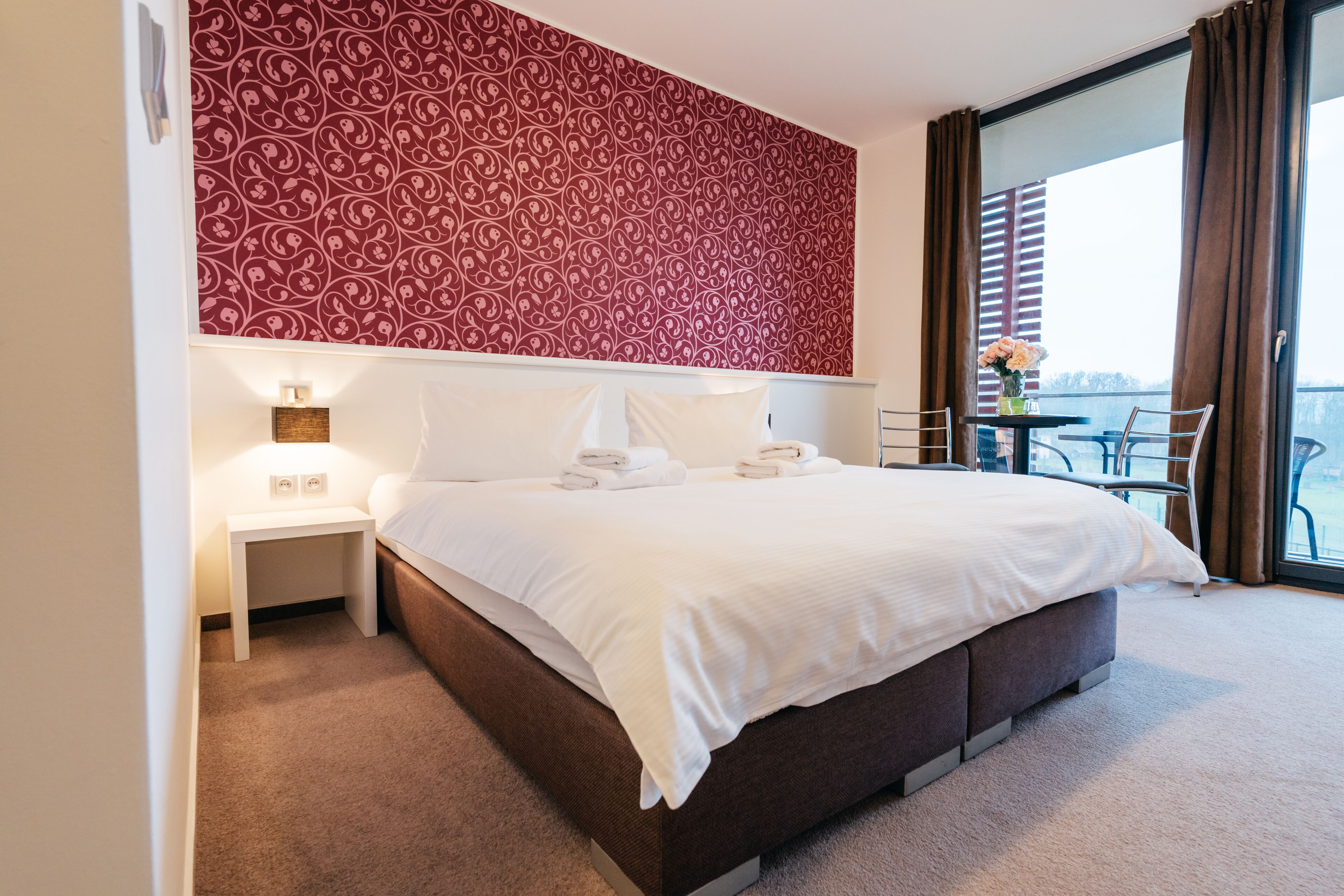 Quarto de hotel com uma cama grande, uma mesa de cabeceira, uma pequena mesa com um vaso de flores e vista para o exterior através de uma janela ampla.