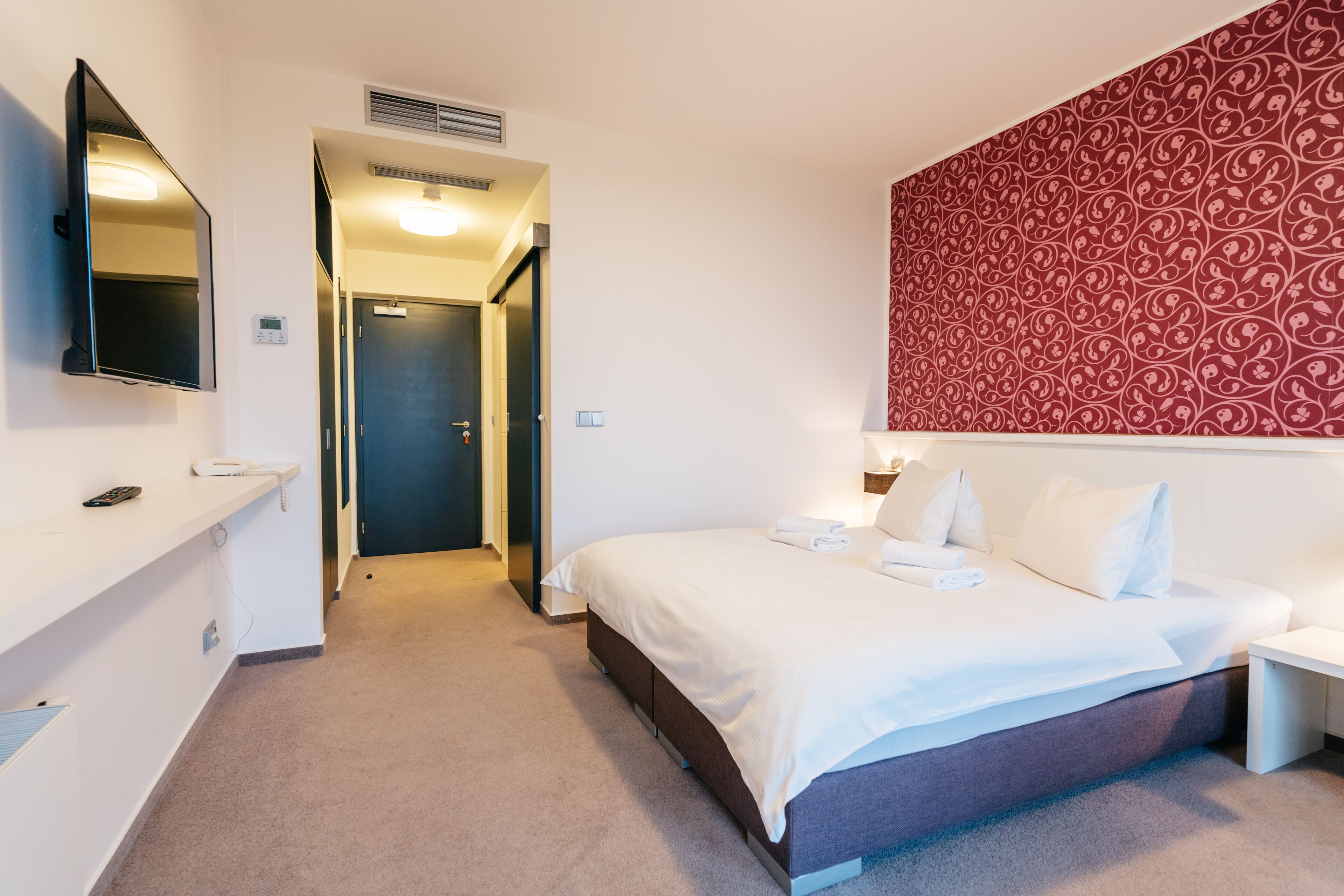 Quarto de hotel com cama, mesa de cabeceira e um espelho grande. Há uma porta que dá para outro cômodo e uma parede com padrão.