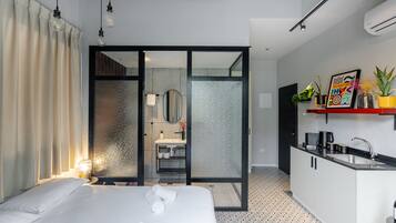 Quarto de hotel moderno com cama, banheiro com porta de vidro, cozinha compacta e pequena sala de estar.
