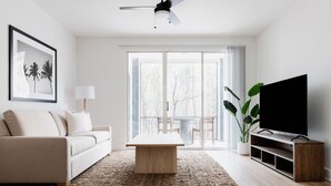 Uma sala de estar moderna com um sofá, uma mesa de centro, uma TV sobre um suporte de madeira e uma janela grande com vista para árvores.