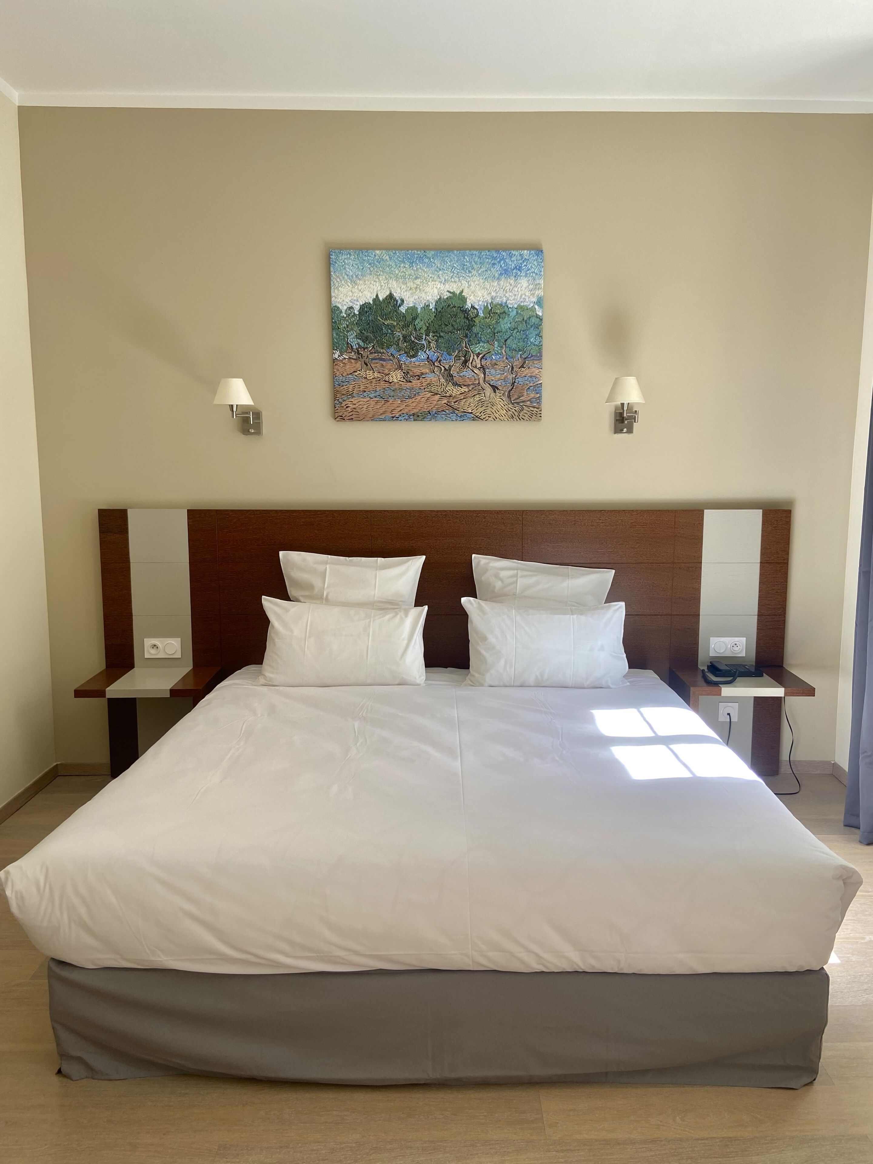 Uma cama grande com lençóis brancos, cabeceira de madeira, duas mesas de cabeceira com abajures e um quadro na parede.