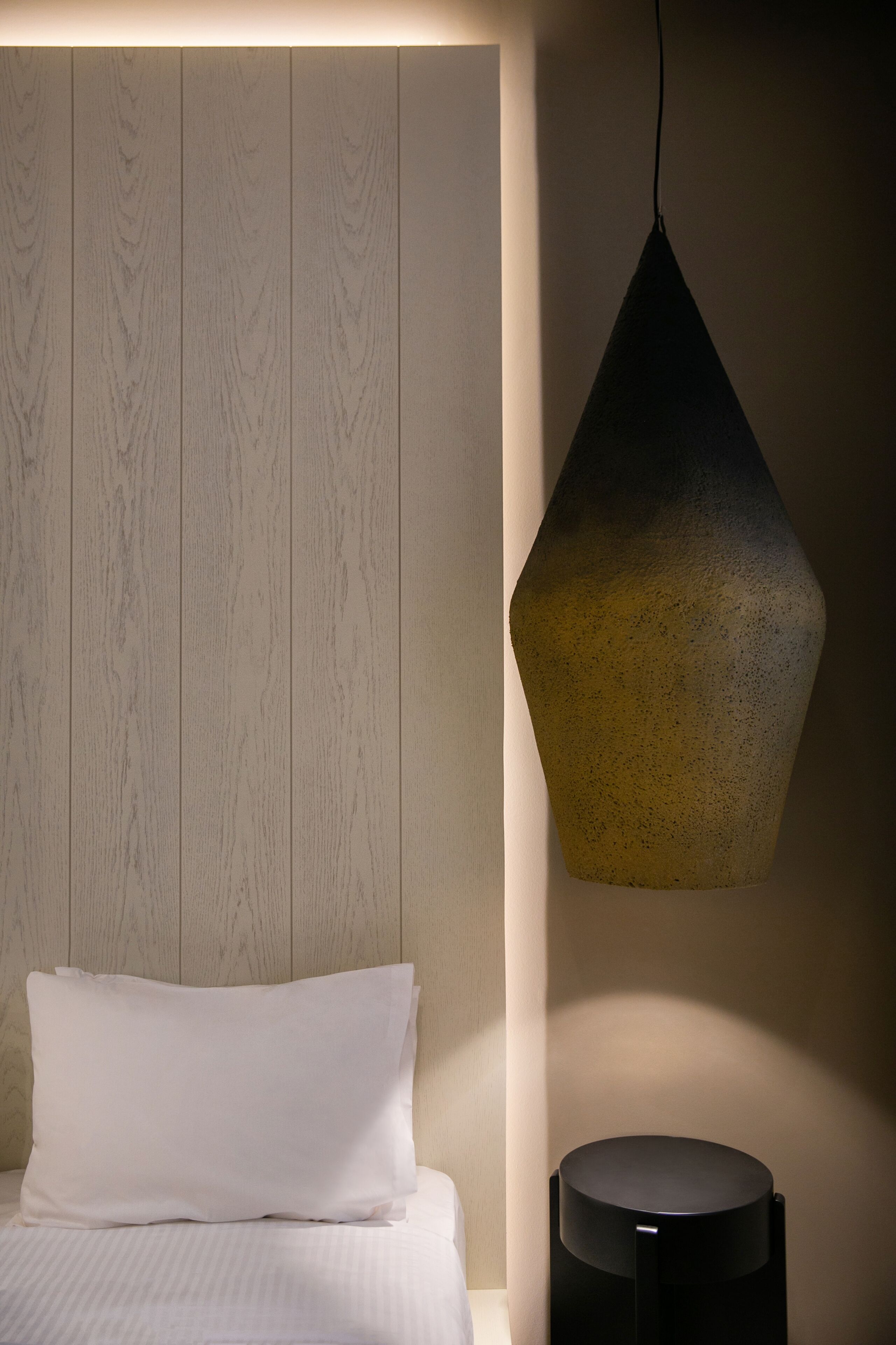 Uma cama com travesseiro branco e mesa de cabeceira preta com abajur.