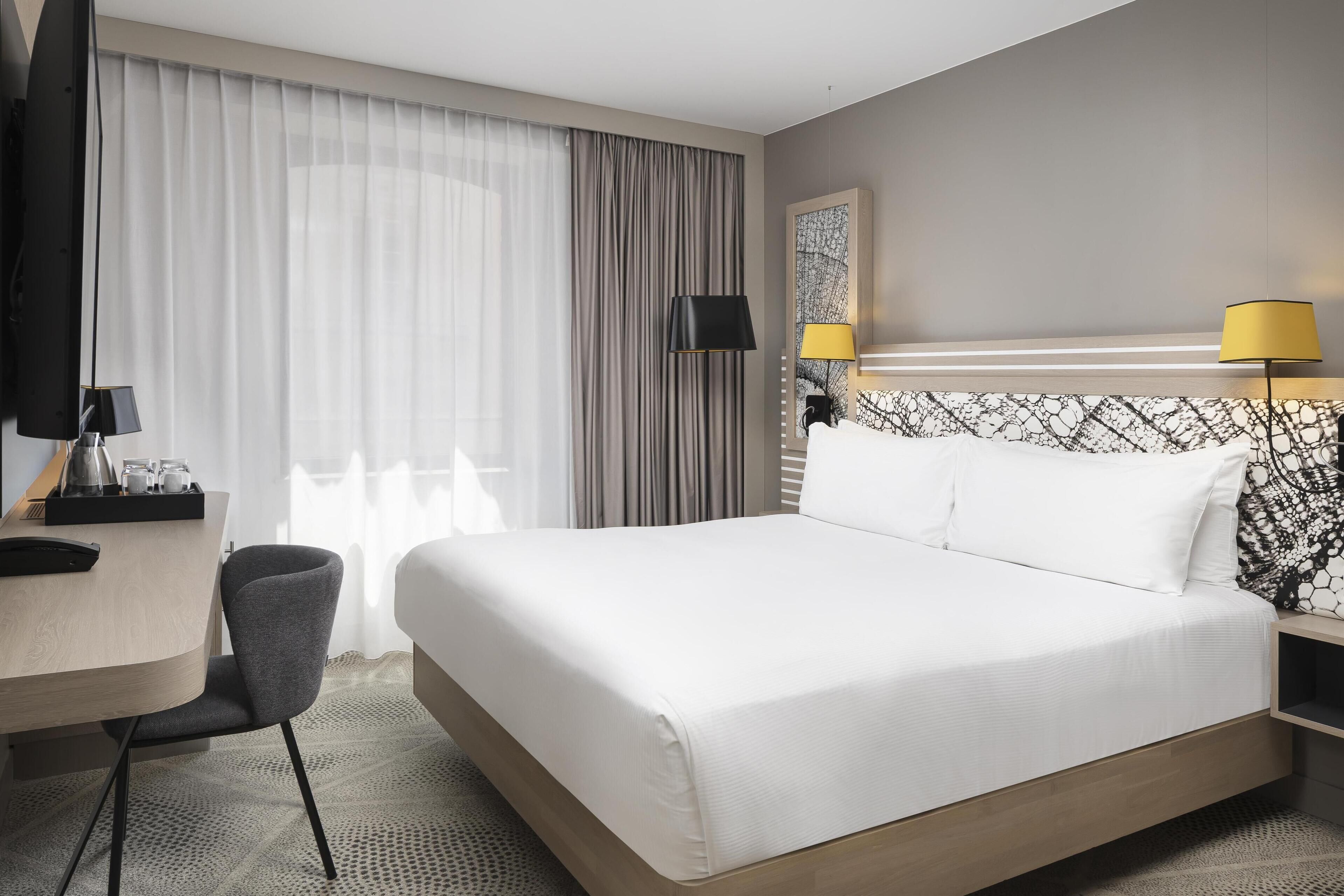 Quarto de hotel moderno com uma cama grande, uma escrivaninha com cadeira, uma televisão e duas luminárias.
