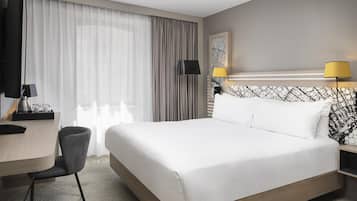 Quarto de hotel moderno com uma cama grande, uma escrivaninha com cadeira, uma televisão e duas luminárias.
