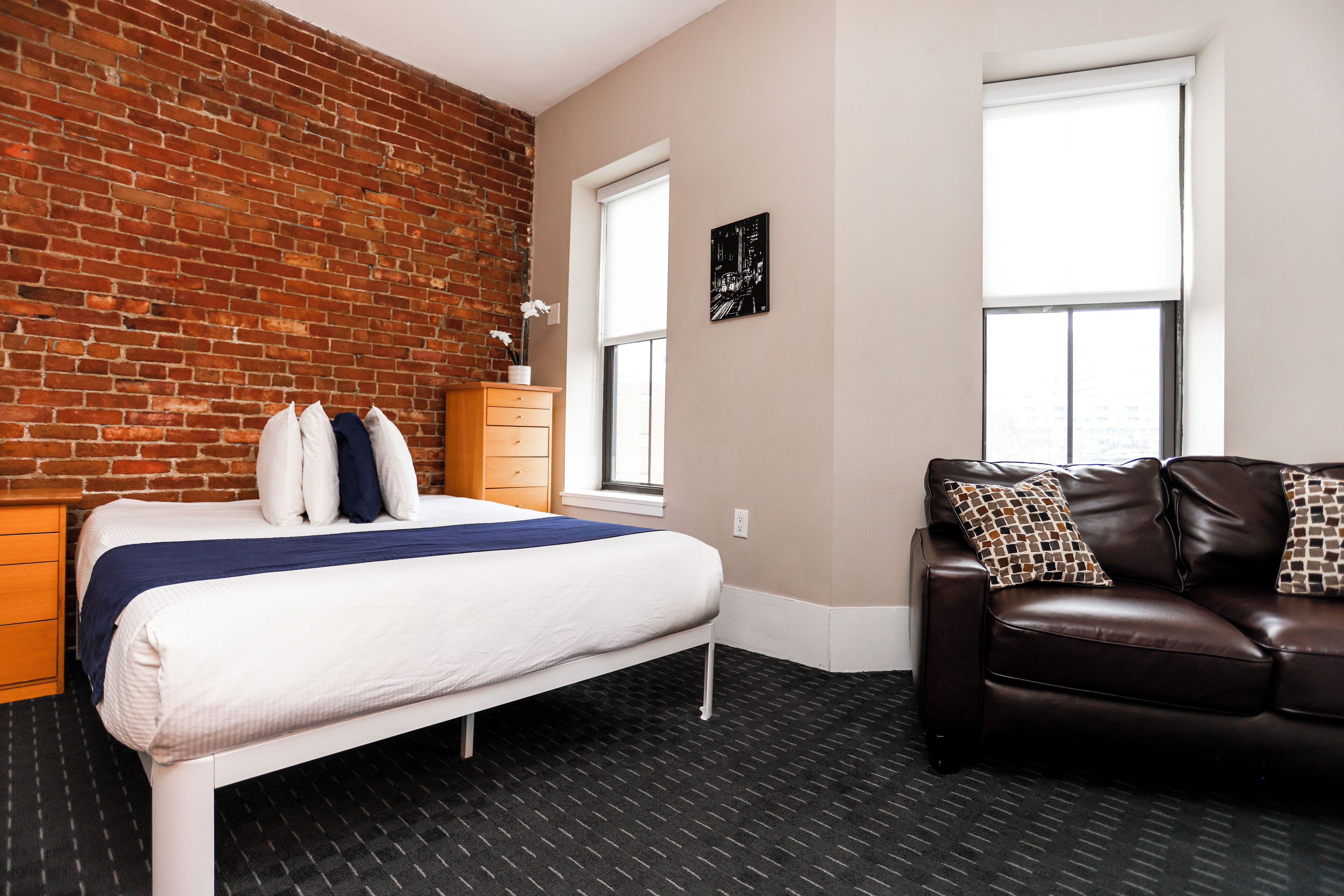 Quarto com cama, cômoda, sofá de couro marrom e parede de tijolos.