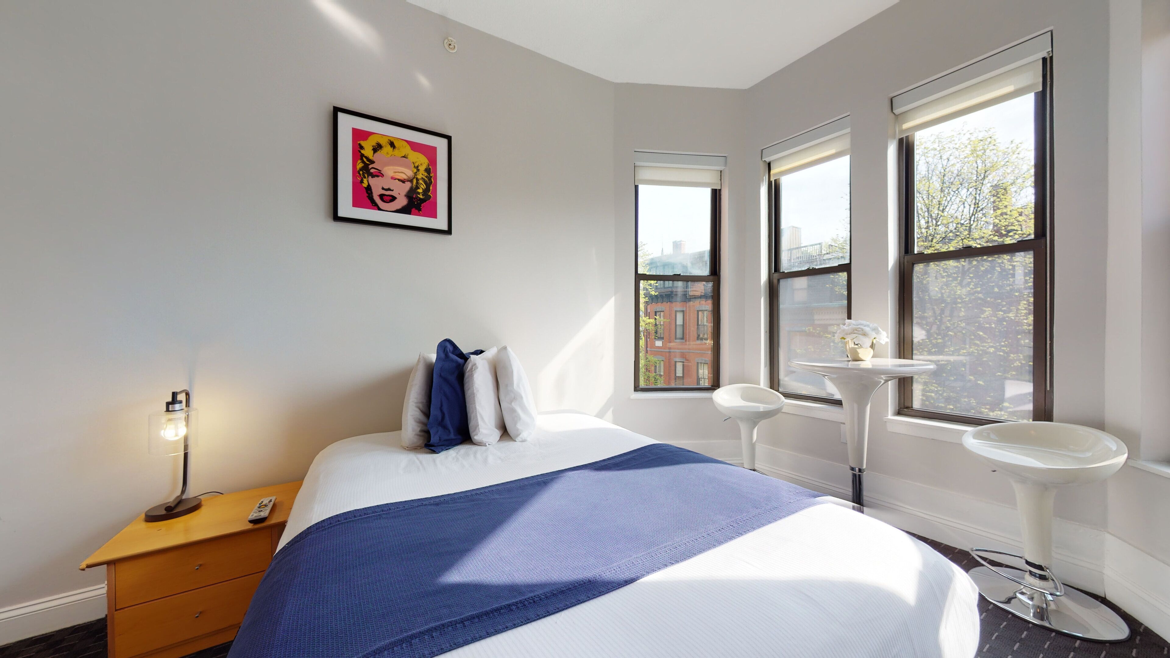 Um quarto moderno com cama, mesa de cabeceira, luminária e janela com vista para edifícios.