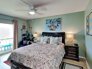 Quarto com cama, duas mesinhas de cabeceira, ventilador de teto, janela com cortinas e vista para o exterior.