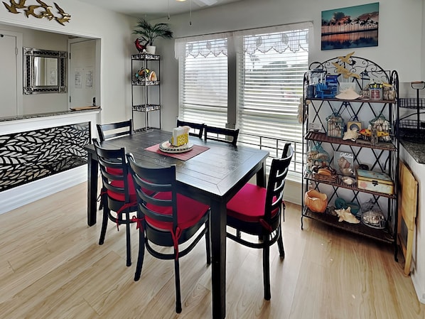 Uma área de refeições com uma mesa e cadeiras, uma estante decorativa com diversos objetos, e uma janela com persianas.